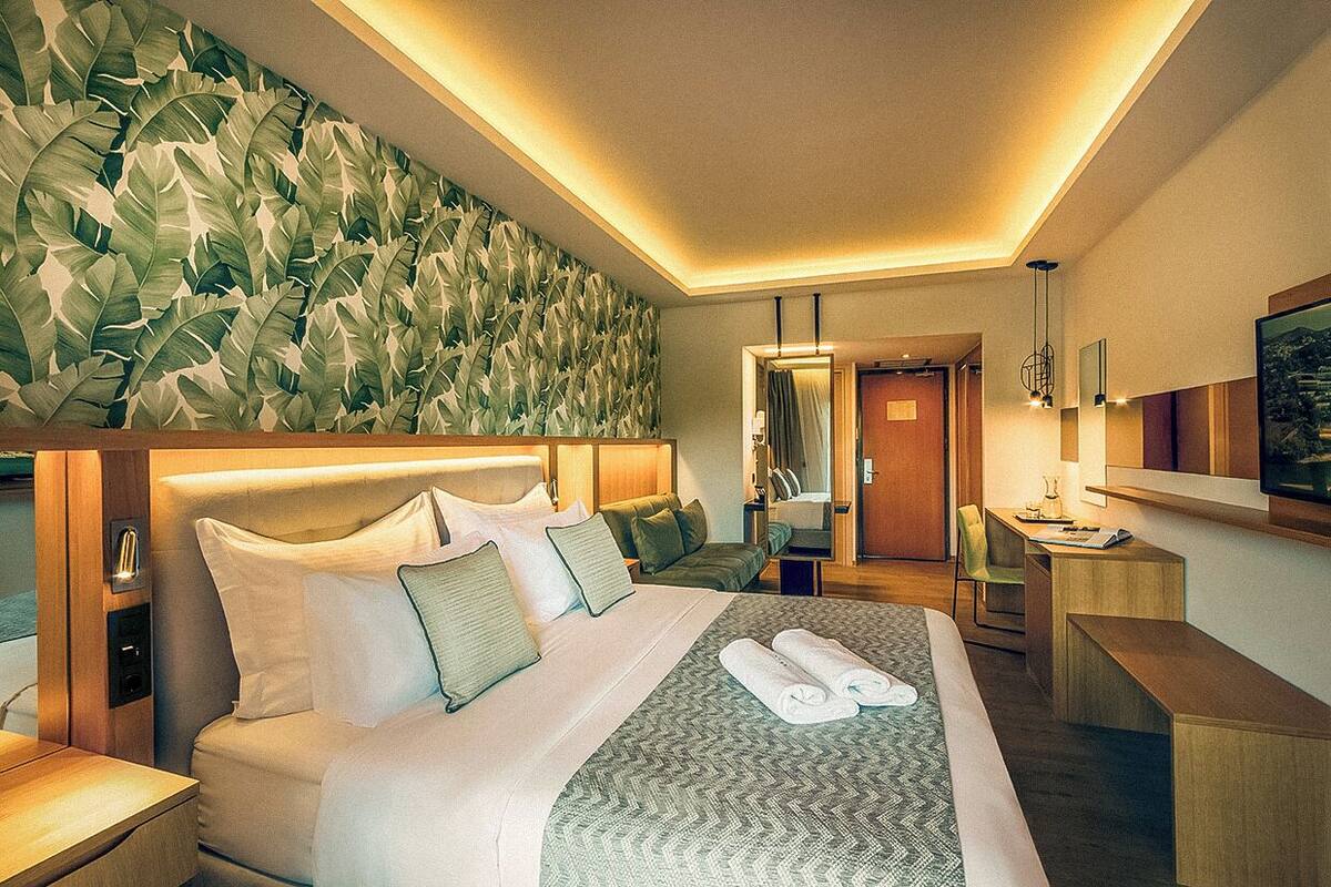 Quarto de hotel com uma cama grande, uma escrivaninha, uma cadeira, um espelho e uma parede com padrão de folhas.