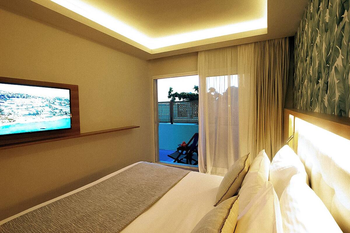 Quarto de hotel com cama, televisão na parede, varanda com cadeira e vista para o exterior.