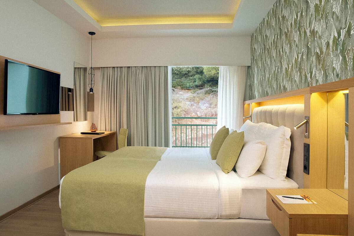 Quarto de hotel com cama, televisão, escrivaninha e varanda com vista para o verde.
