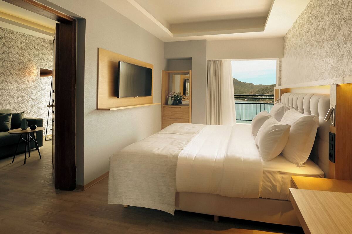 Quarto moderno com uma cama grande, televisão na parede, varanda com vista para o mar e escrivaninha de madeira.
