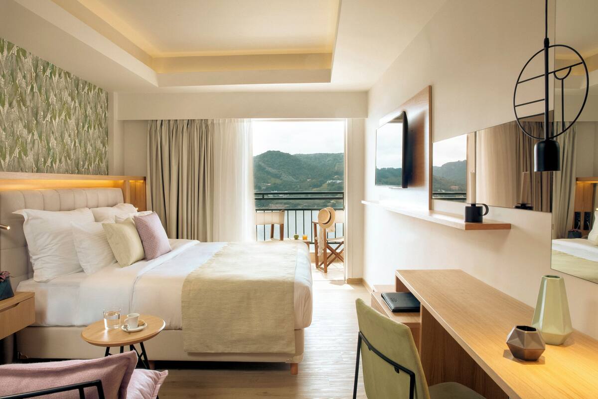 Quarto de hotel com uma cama grande, uma mesa de escritório, uma cadeira, uma mesinha, uma TV e uma varanda com vista para as colinas.