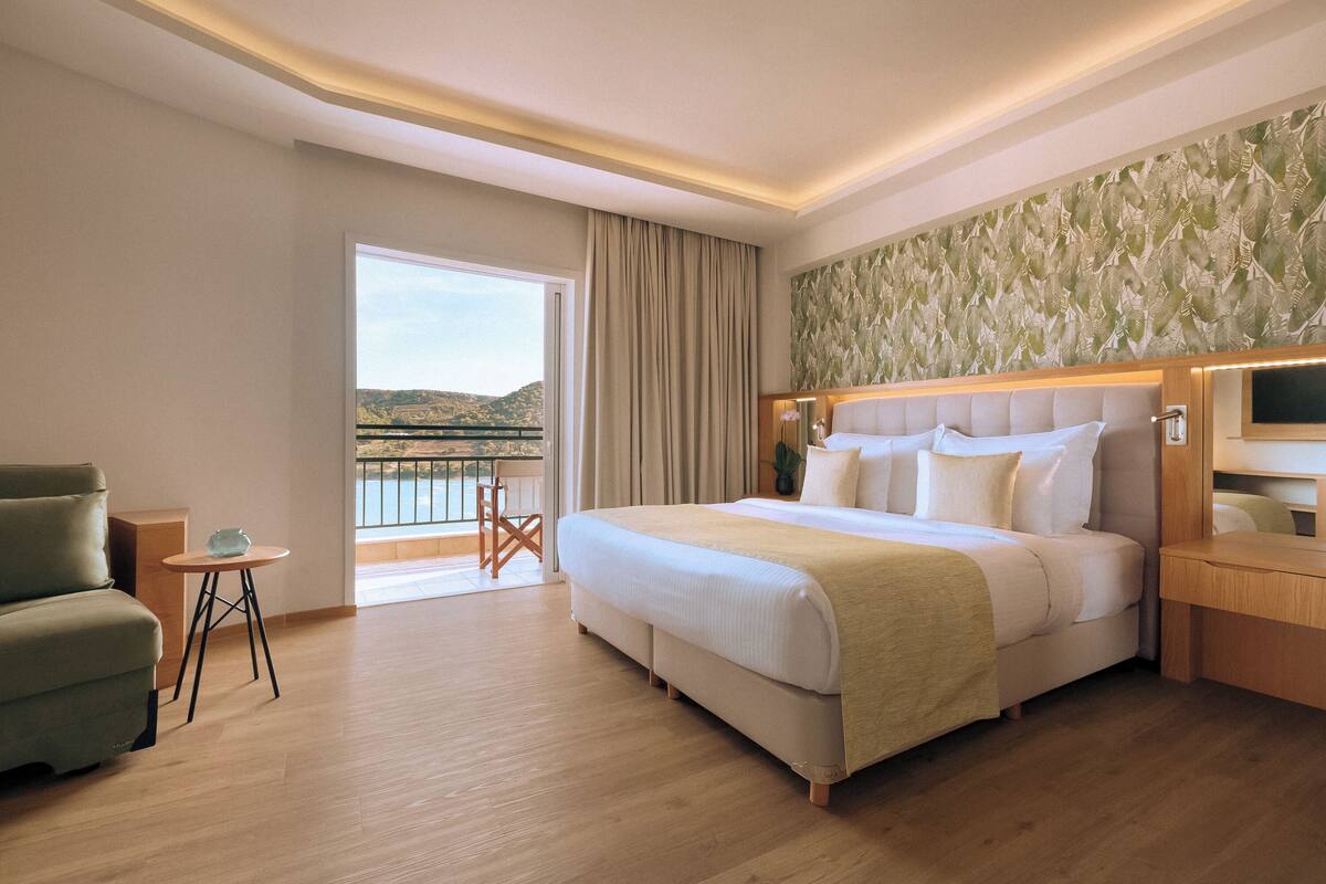 Quarto de hotel com uma cama grande, uma cadeira, uma mesinha e varanda com vista para as colinas.