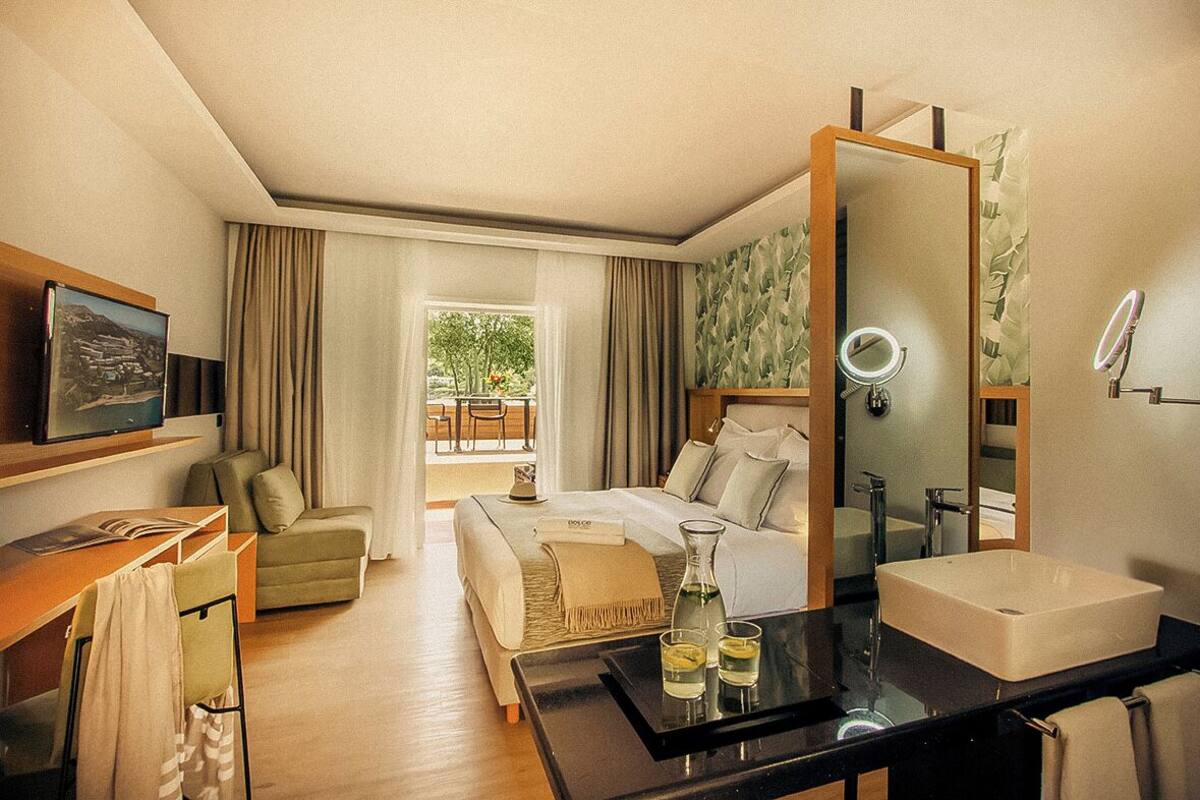 Quarto de hotel com cama, escrivaninha, cadeira, espelho grande, pia e vista para o exterior.
