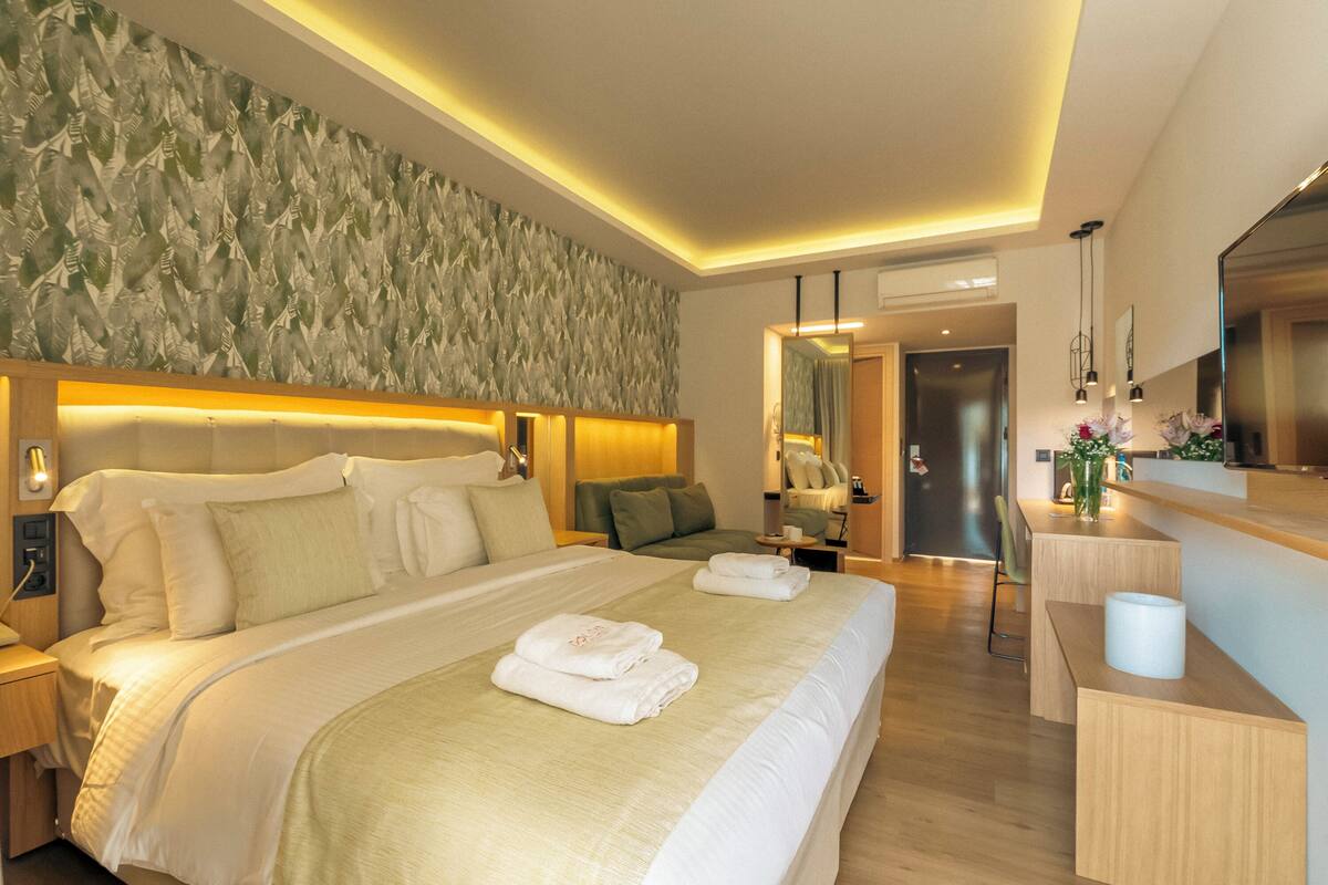 Quarto de hotel moderno com uma cama grande, mesa de cabeceira, espelho e um pequeno espaço de estar com uma poltrona e uma mesa.