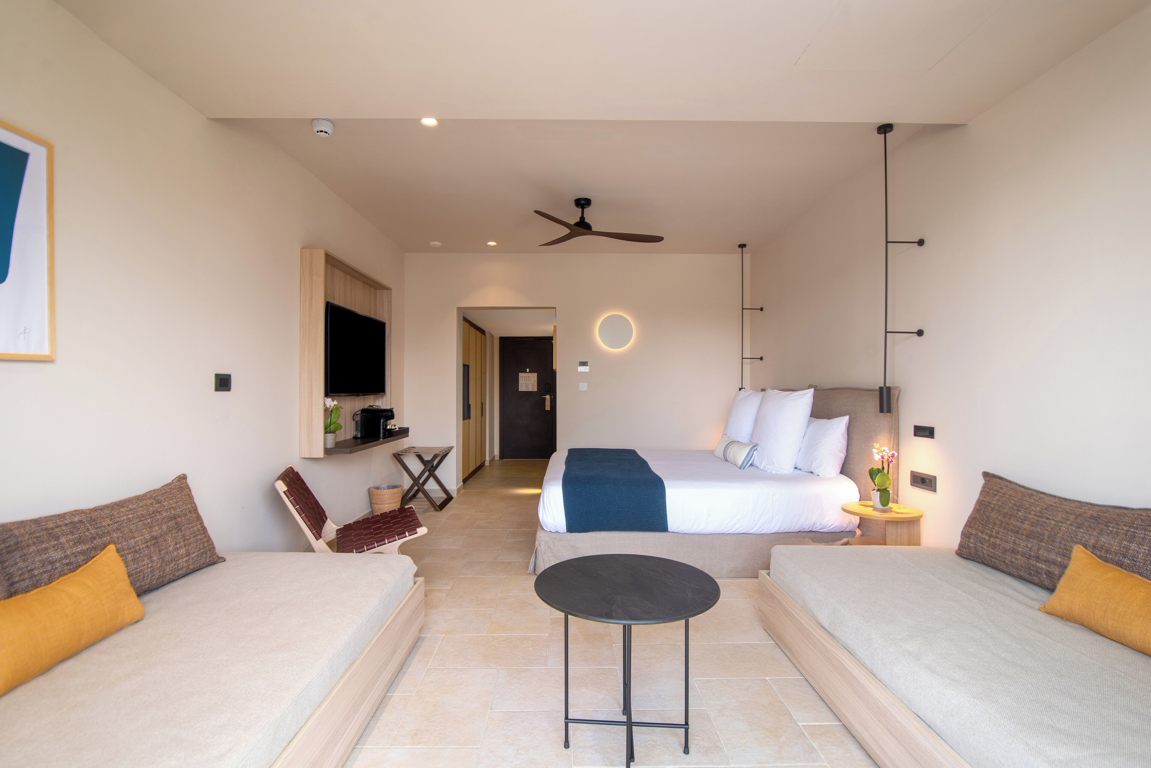 Quarto de hotel moderno com uma cama grande, dois sofás, uma pequena mesa e um ventilador de teto.