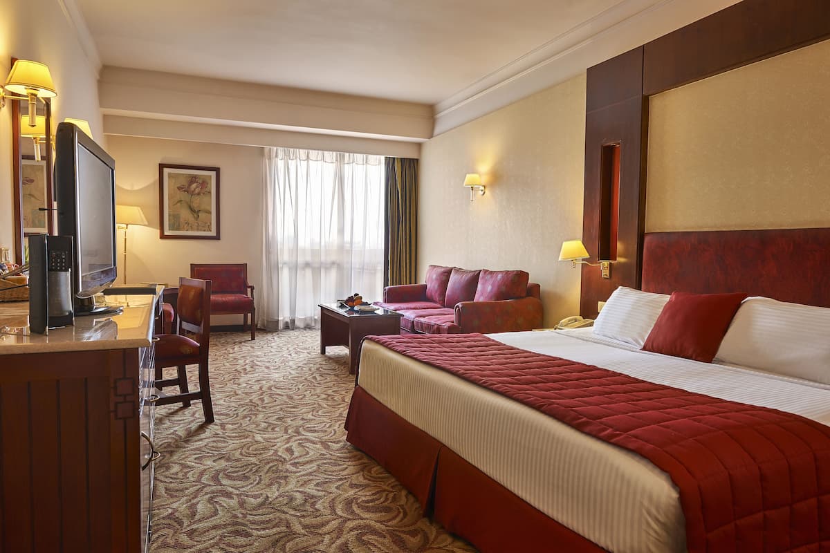 Quarto de hotel com uma cama grande, uma escrivaninha com computador, uma cadeira, um sofá e uma televisão.