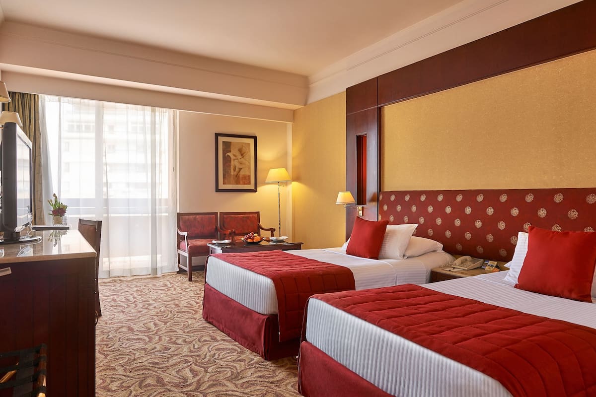 Quarto de hotel com duas camas, uma escrivaninha, uma cadeira, um abajur e uma janela com cortinas.
