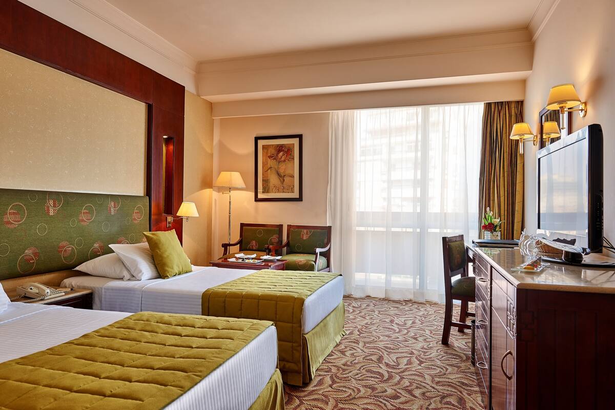 Quarto de hotel com duas camas, uma mesa com televisão, uma cadeira e uma luminária.