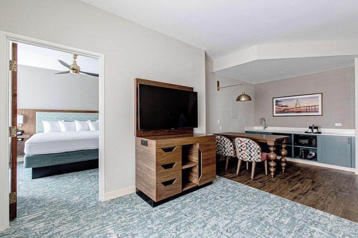 Quarto com cama, TV sobre apoio de madeira, área de refeições com cadeiras e cozinha com pia e armários.