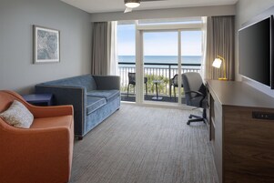 Quarto de hotel com sofá, poltrona, televisão e varanda com vista para o mar.