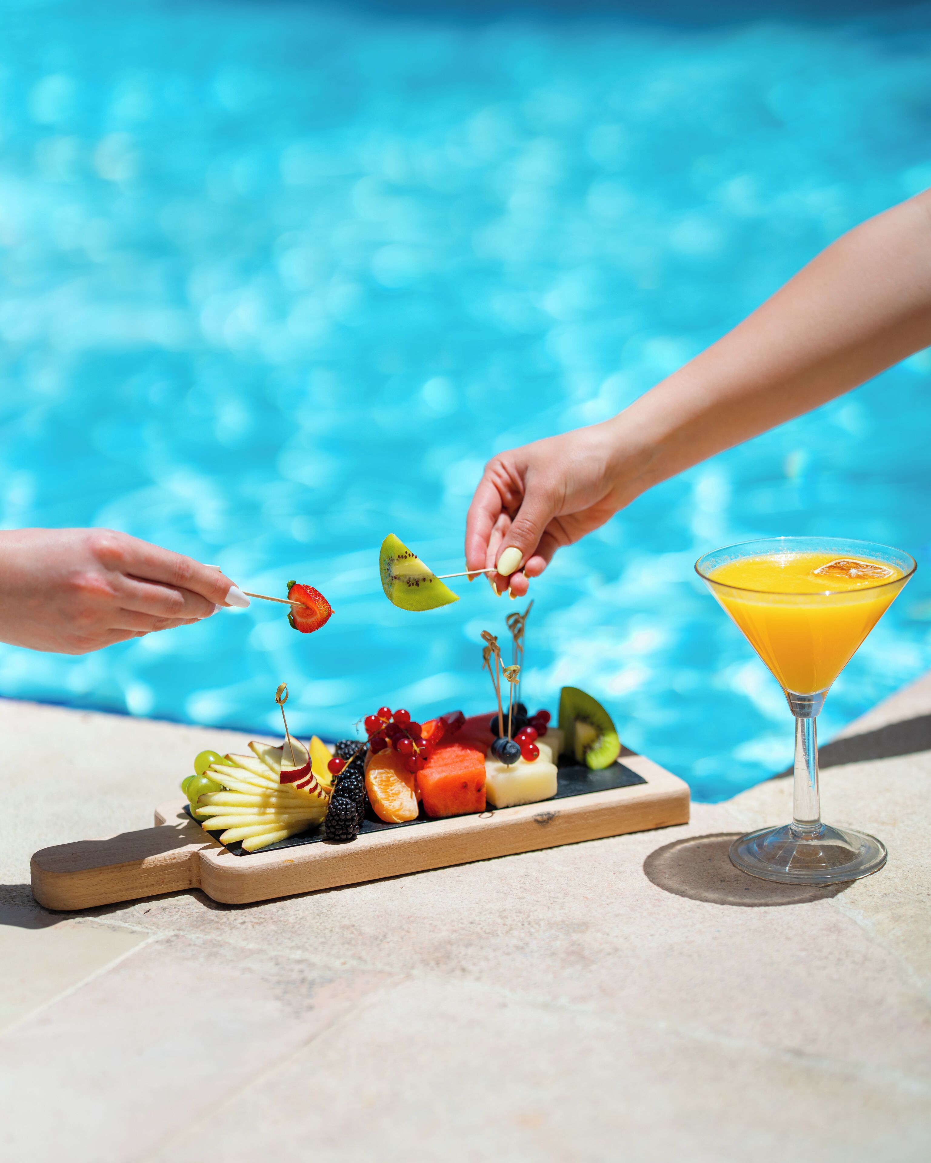 Uma tábua de madeira com diversas frutas e um copo de martini com bebida de laranja à beira de uma piscina.