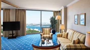 Quarto de hotel com uma janela grande, um sofá, uma poltrona, uma mesa de centro e uma televisão.