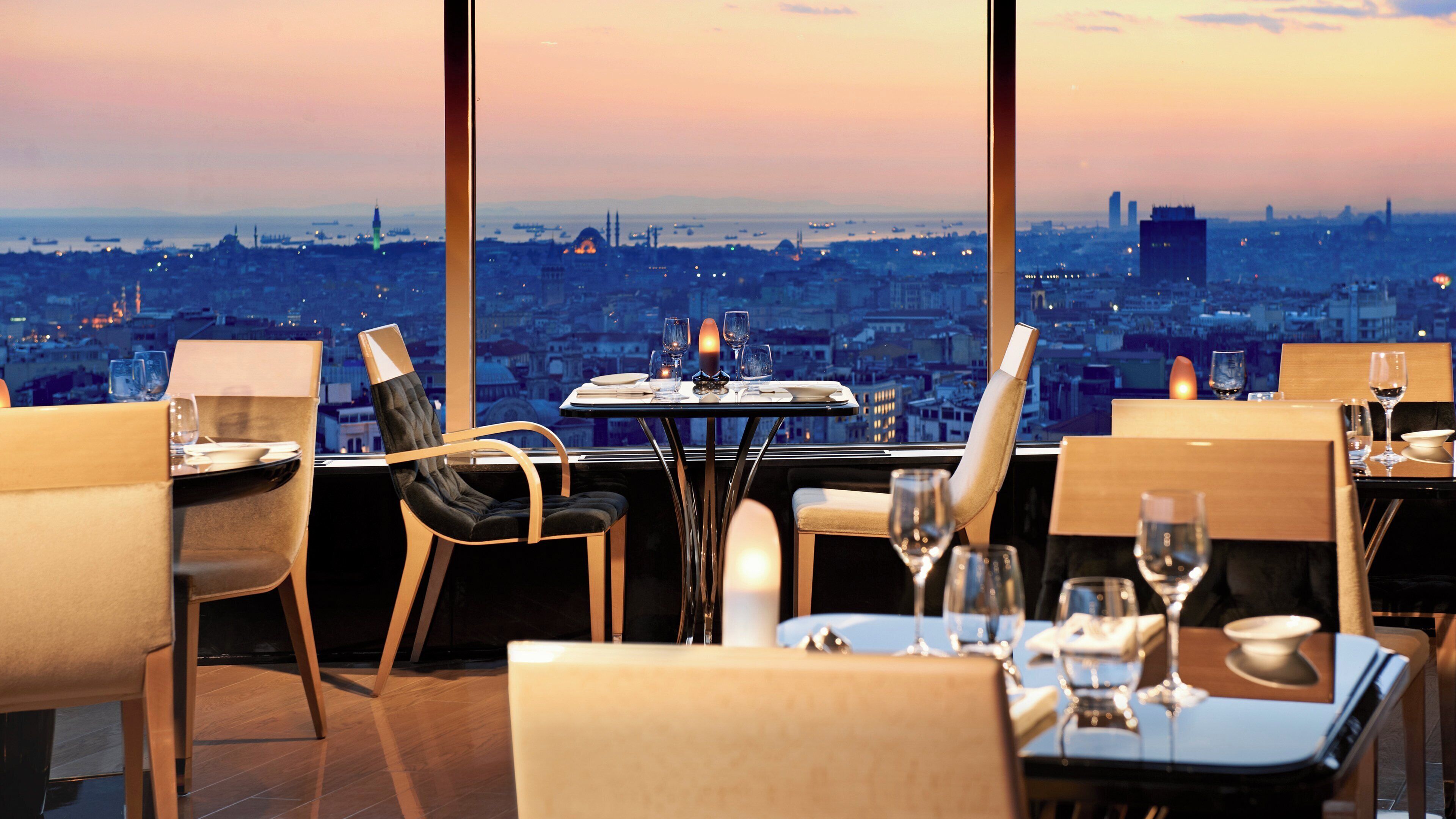 Espaço de refeições com mesa posta para dois, com vista para a paisagem urbana ao entardecer.