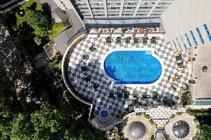 Vista aérea de um hotel com piscina central, área de estar com padrão xadrez e vegetação ao redor.
