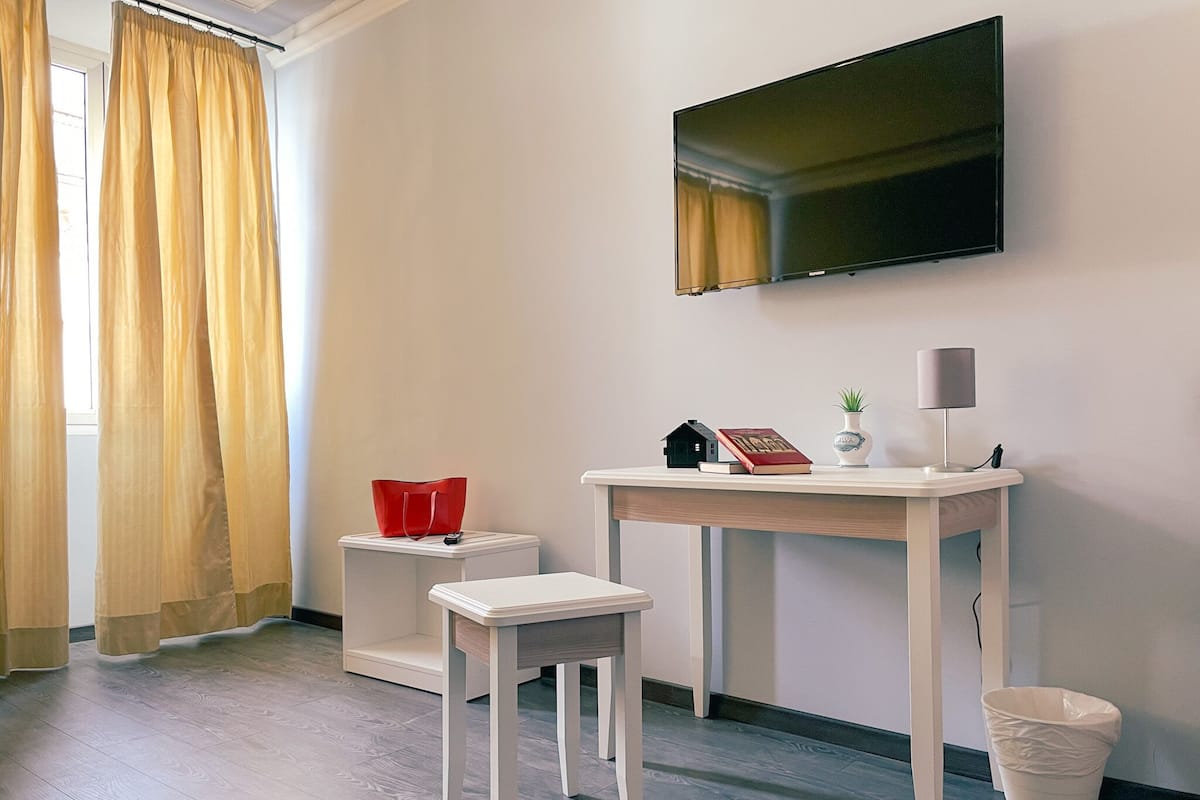 Quarto de hotel com televisão na parede, uma mesa pequena com luminária, lixeira e janela com cortinas.