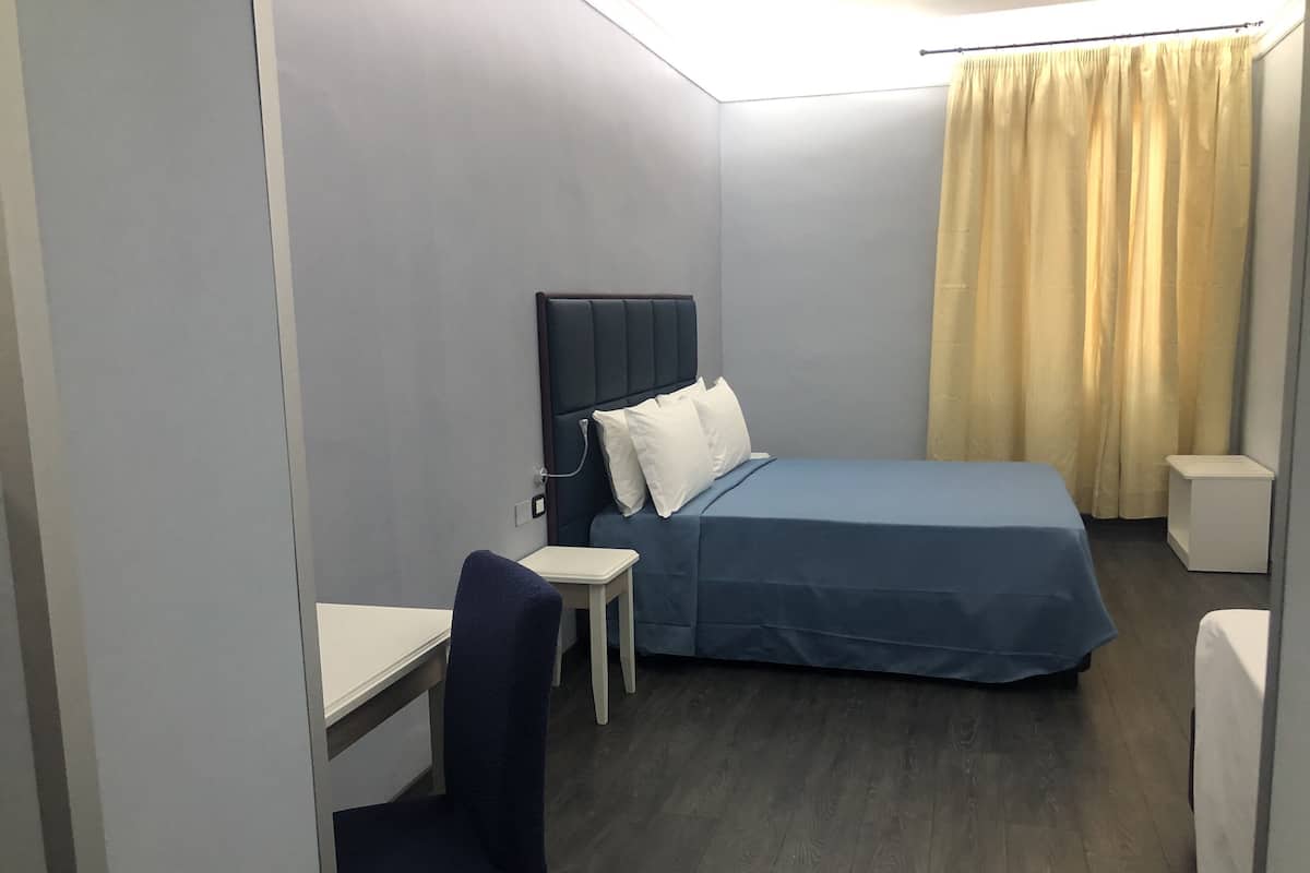 Quarto de hotel pequeno, com uma cama, escrivaninha, cadeira e mesa de cabeceira.