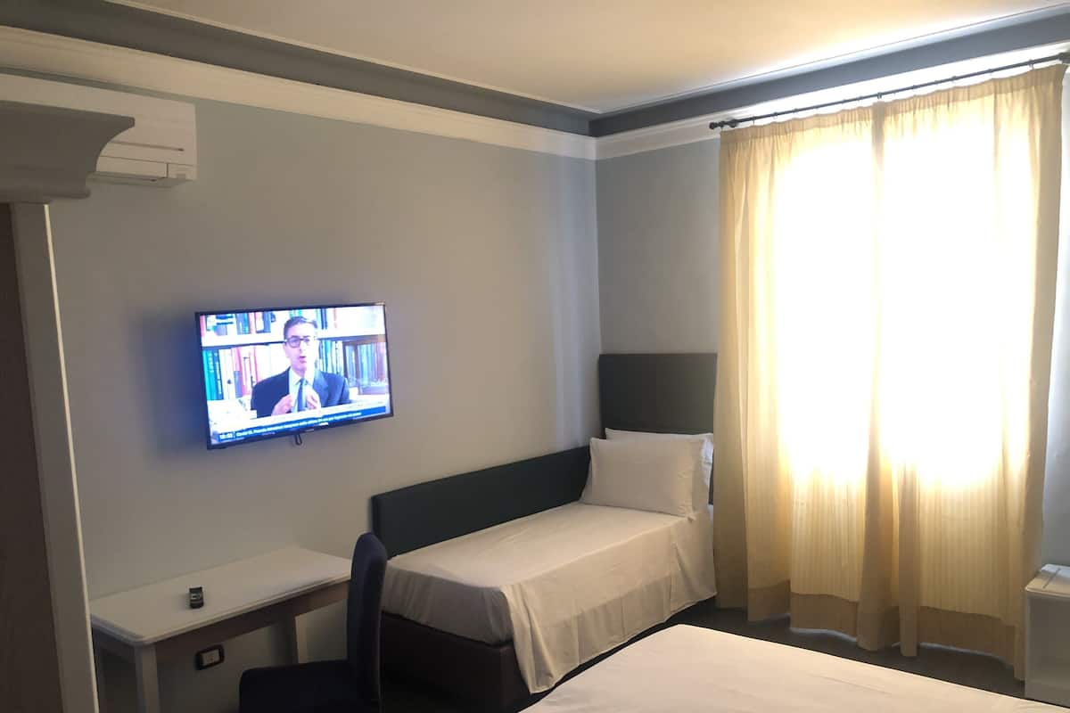 Quarto de hotel com cama, escrivaninha, cadeira, televisão e janela com cortinas.