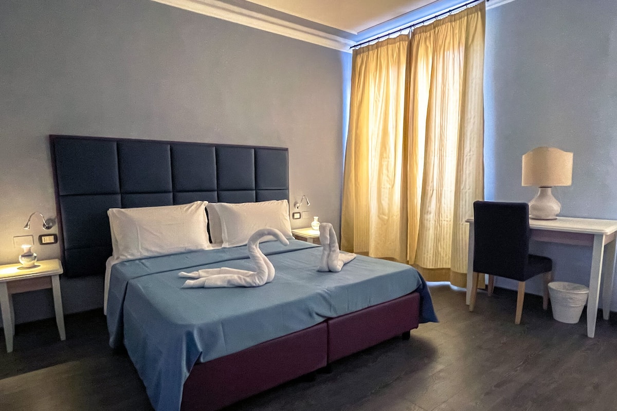 Quarto de hotel com cama, escrivaninha, cadeira e janela com cortinas.