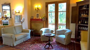 Uma sala de estar acolhedora, com duas poltronas, uma mesa pequena, uma lareira e uma estante com garrafas de vinho.