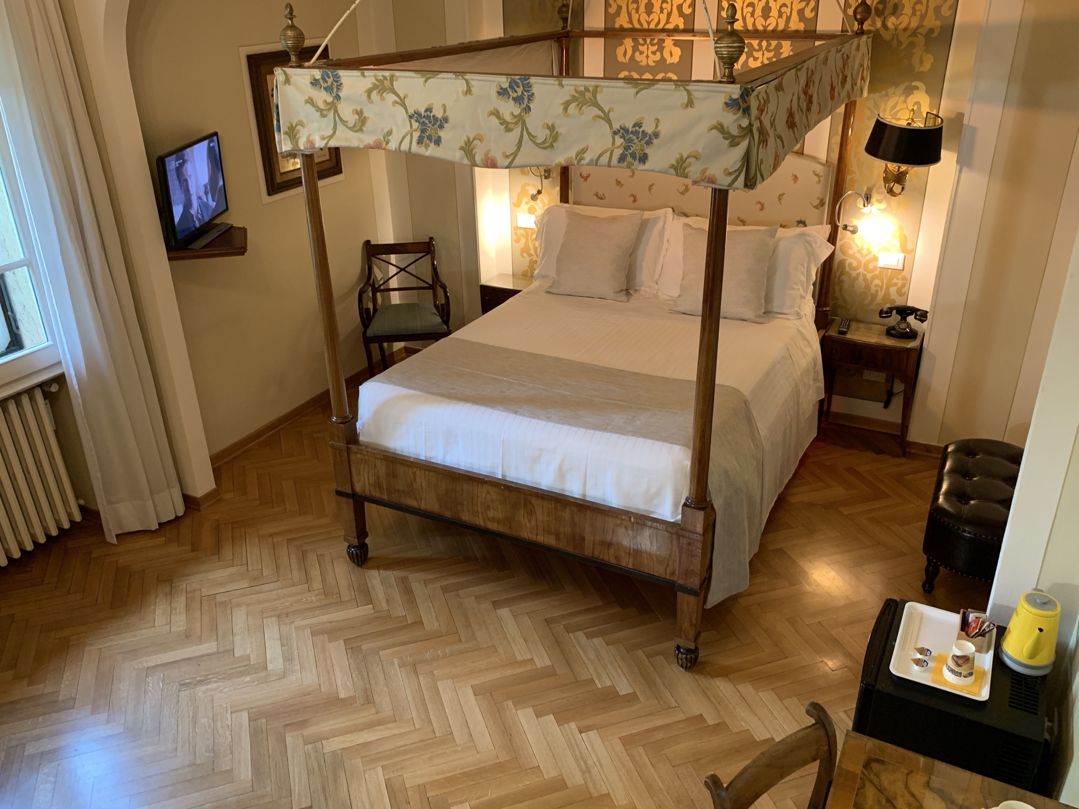 Uma cama com dossel adornada com detalhes florais, uma mesinha de cabeceira com abajur, uma cadeira e uma pequena mesa com uma bandeja de objetos.