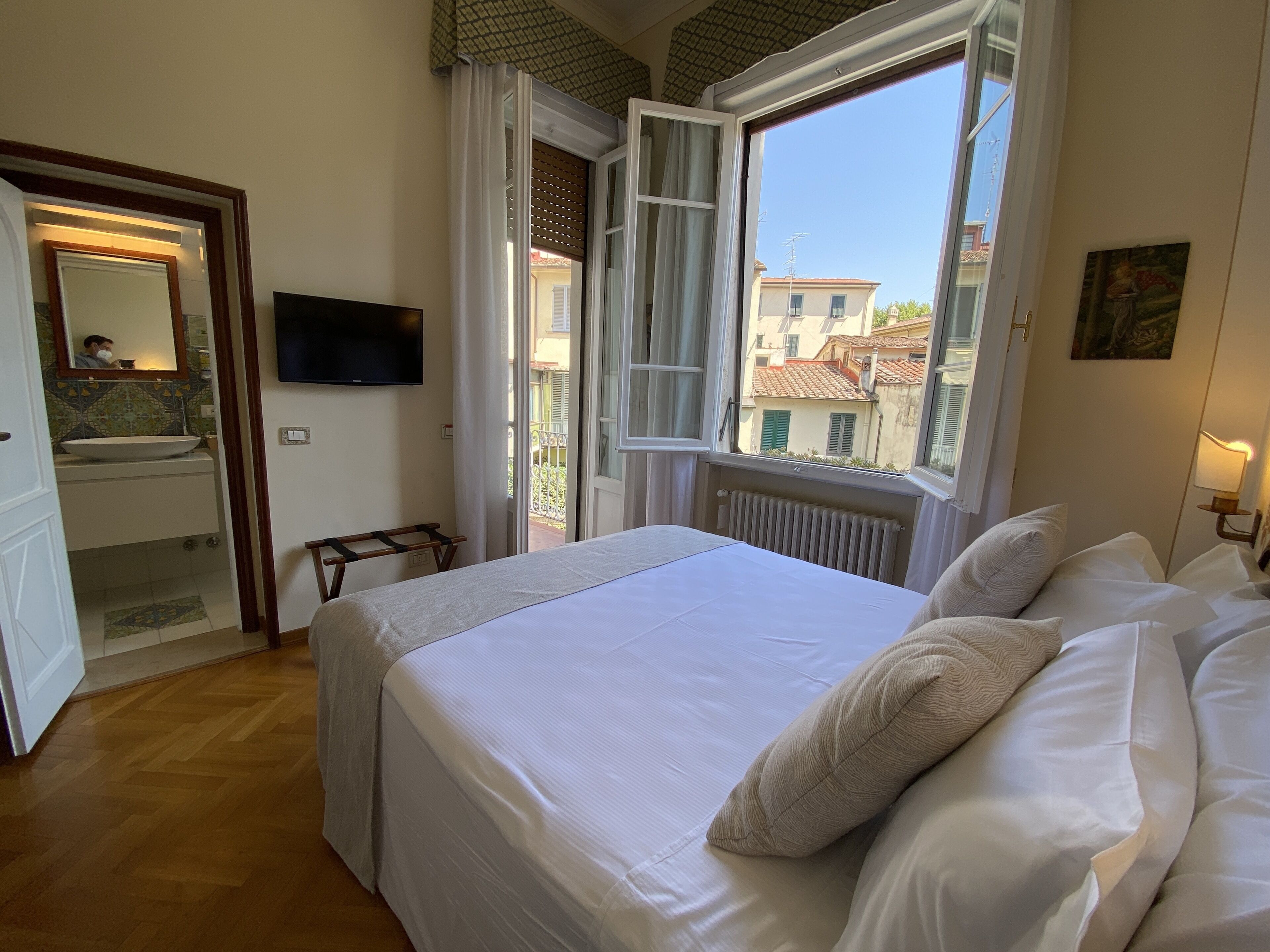 Quarto com cama grande, televisão, banheiro com pia e vista para edifícios por uma janela aberta.