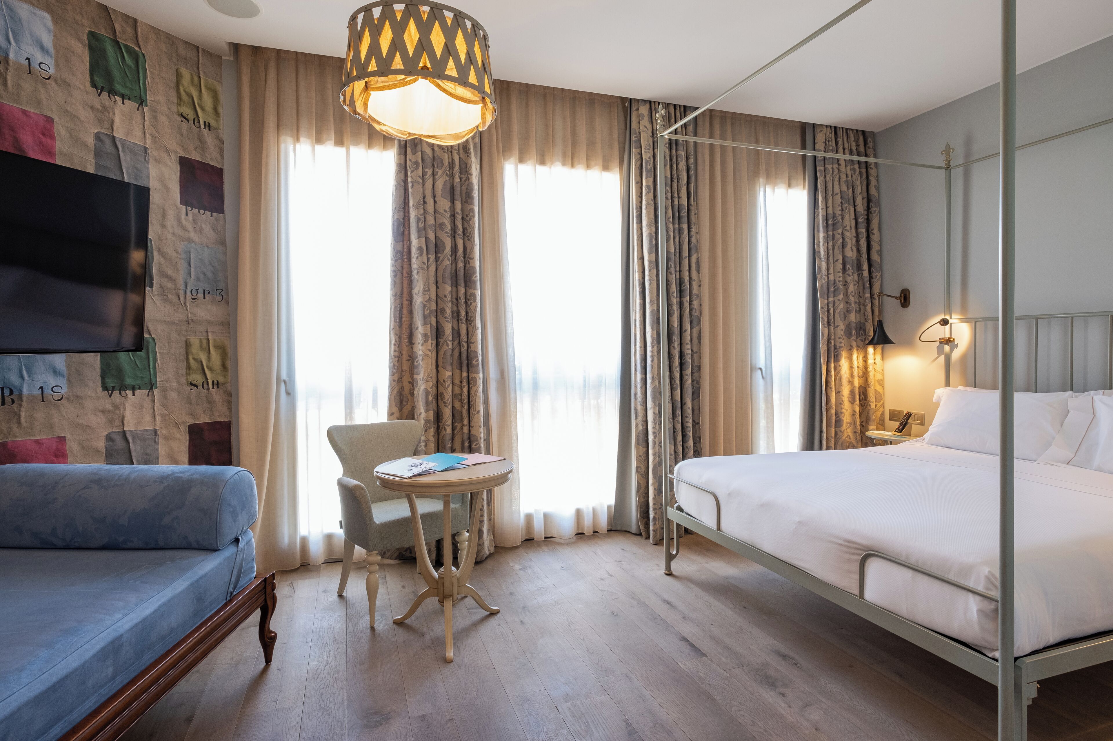 Quarto de hotel com cama, sofá, mesinha com cadeira, televisão na parede e uma janela grande com cortinas.