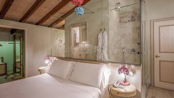 Quarto de hotel com uma cama grande, mesa de cabeceira com luminária, espelho e área de chuveiro.