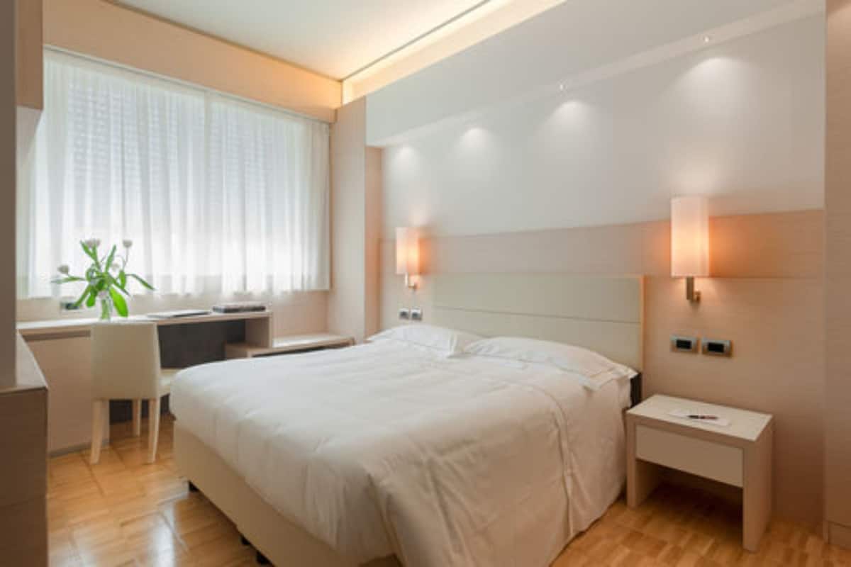 Quarto moderno com uma cama grande, uma escrivaninha e uma cadeira. Há uma janela com cortinas e uma planta na escrivaninha.
