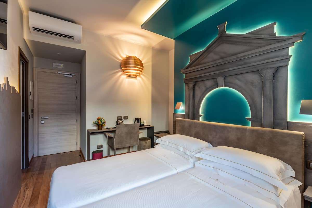 Quarto de hotel moderno com uma cama grande, uma escrivaninha com cadeira e uma parede decorativa com luminária única.