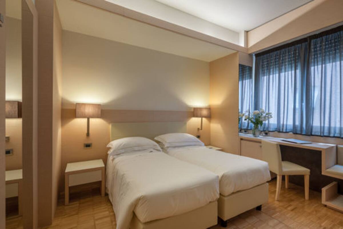 Quarto de hotel com duas camas, uma escrivaninha e uma janela com cortinas.