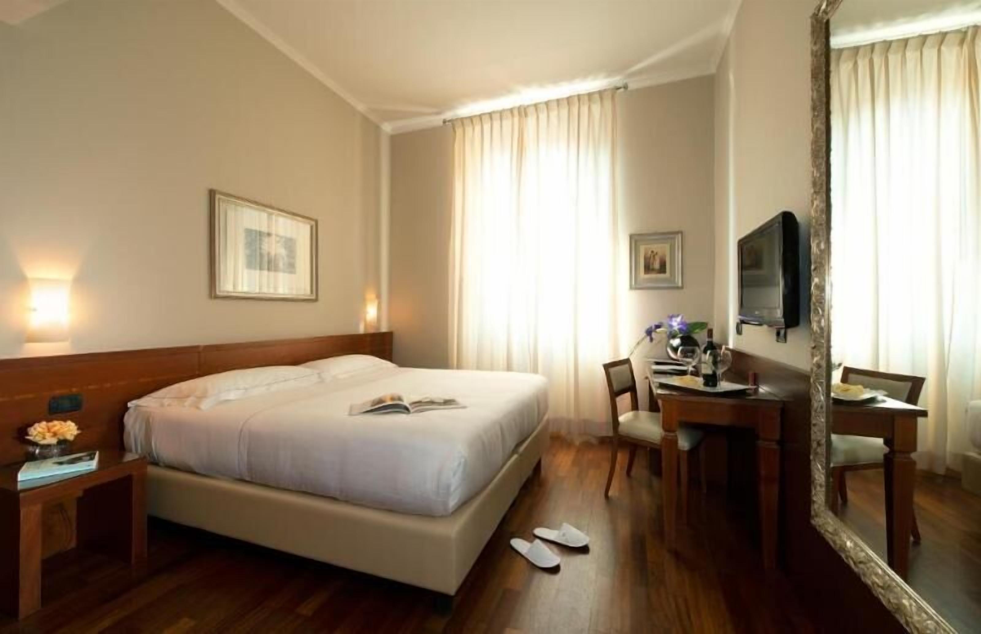 Quarto de hotel com cama, mesa de cabeceira, abajur, escrivaninha, cadeira, televisão e cortinas.
