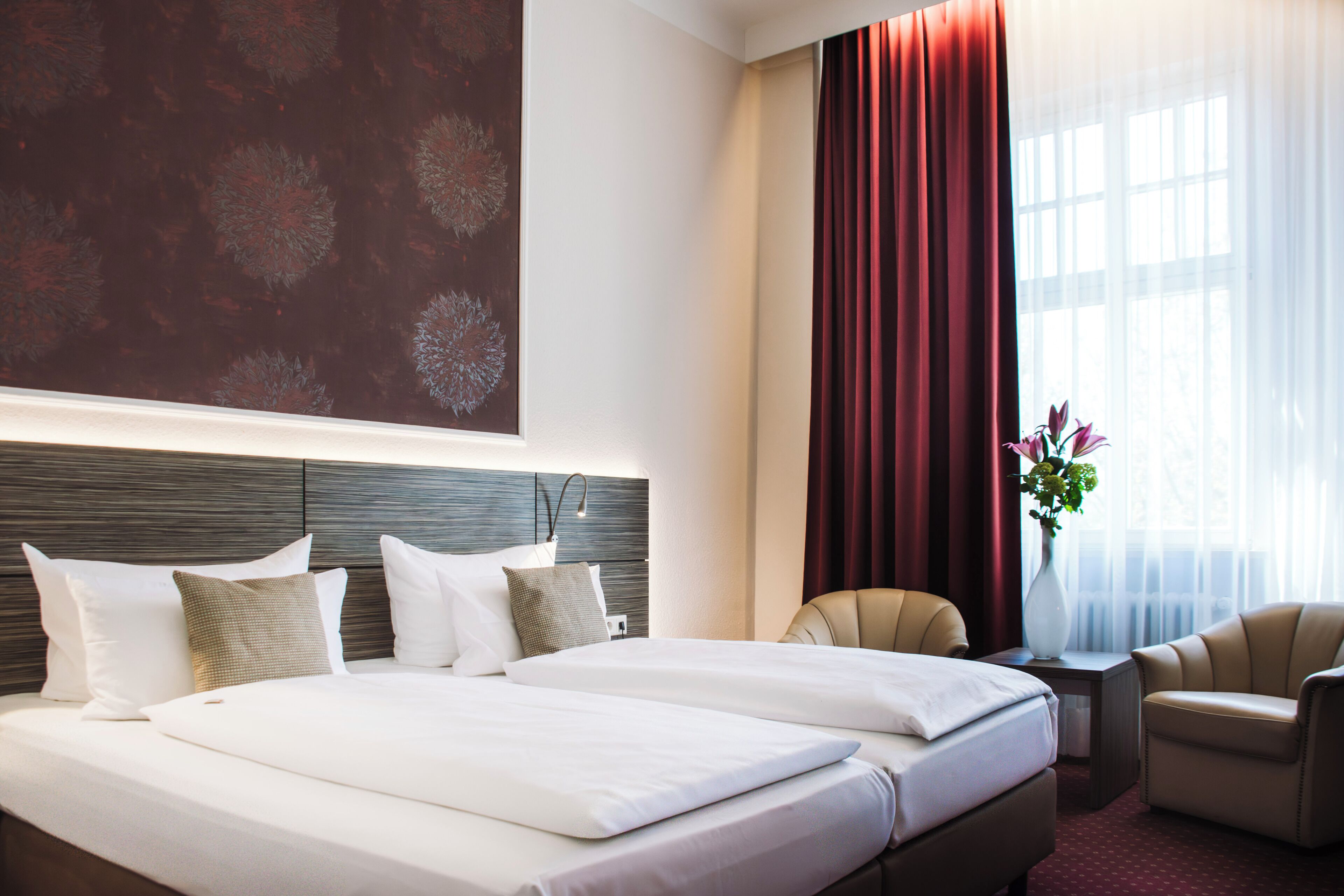 Quarto de hotel com duas camas, uma pintura abstrata grande, um vaso com flores e vista para o exterior.