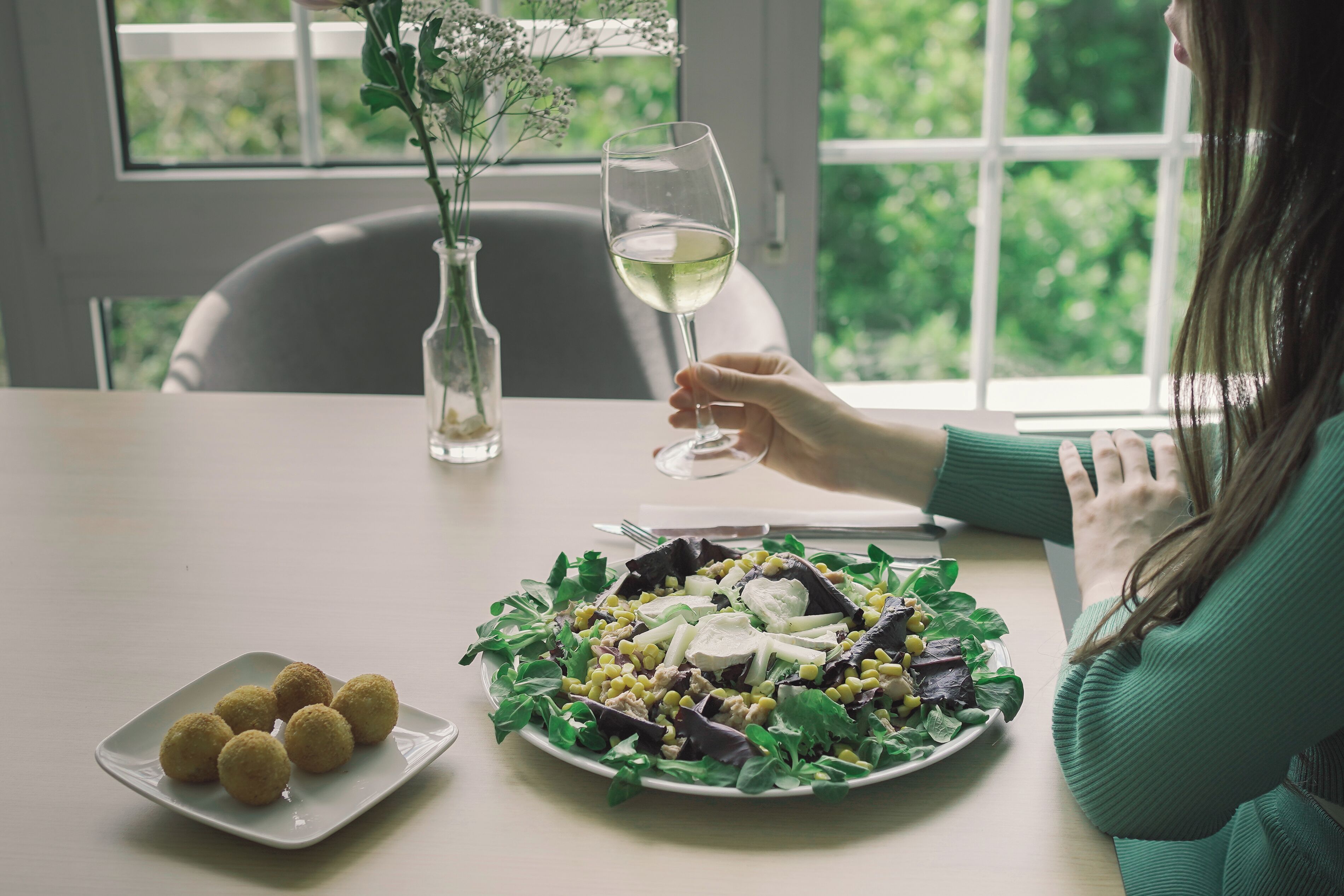 Uma mesa de jantar com uma salada e croquetes, um copo de vinho branco e um vaso com flores.