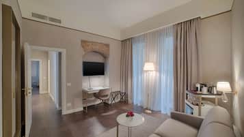 Quarto de hotel com sofá, mesa redonda pequena com um vaso, escrivaninha com cadeira, televisão e área de kitchenette.