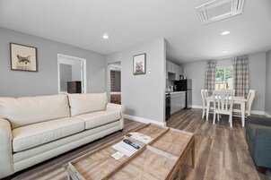 Uma sala de estar moderna com um sofá bege, mesa de centro de madeira e uma área de jantar com mesa e cadeiras brancas.