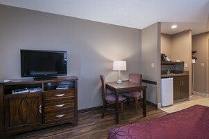 Quarto de hotel com televisão, escrivaninha, cadeira, luminária, minibar e cozinha compacta.