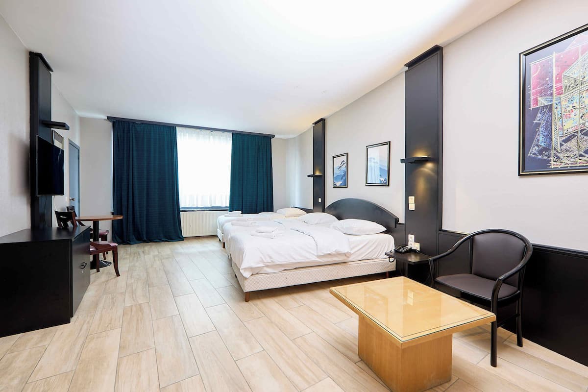 Quarto de hotel com cama, escrivaninha, cadeira, mesa de centro e uma janela grande com cortinas.
