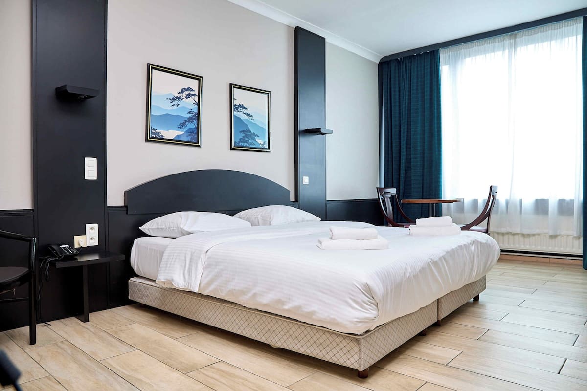 Quarto de hotel moderno com uma cama grande, uma escrivaninha e uma cadeira. Há duas imagens emolduradas na parede, uma janela com cortinas e uma porta com interruptor de luz.