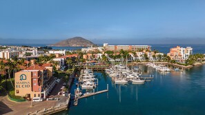 Uma marina com barcos atracados, um hotel chamado 'MARINA EL CID' com uma placa visível, e uma montanha ao fundo.