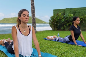 Duas pessoas em tapetes de yoga ao ar livre, com um corpo d'água e vegetação ao fundo.