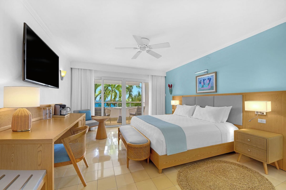 Quarto de hotel com cama, escrivaninha com cadeira, televisão, ventoinha no teto e vista para palmeiras pela janela.