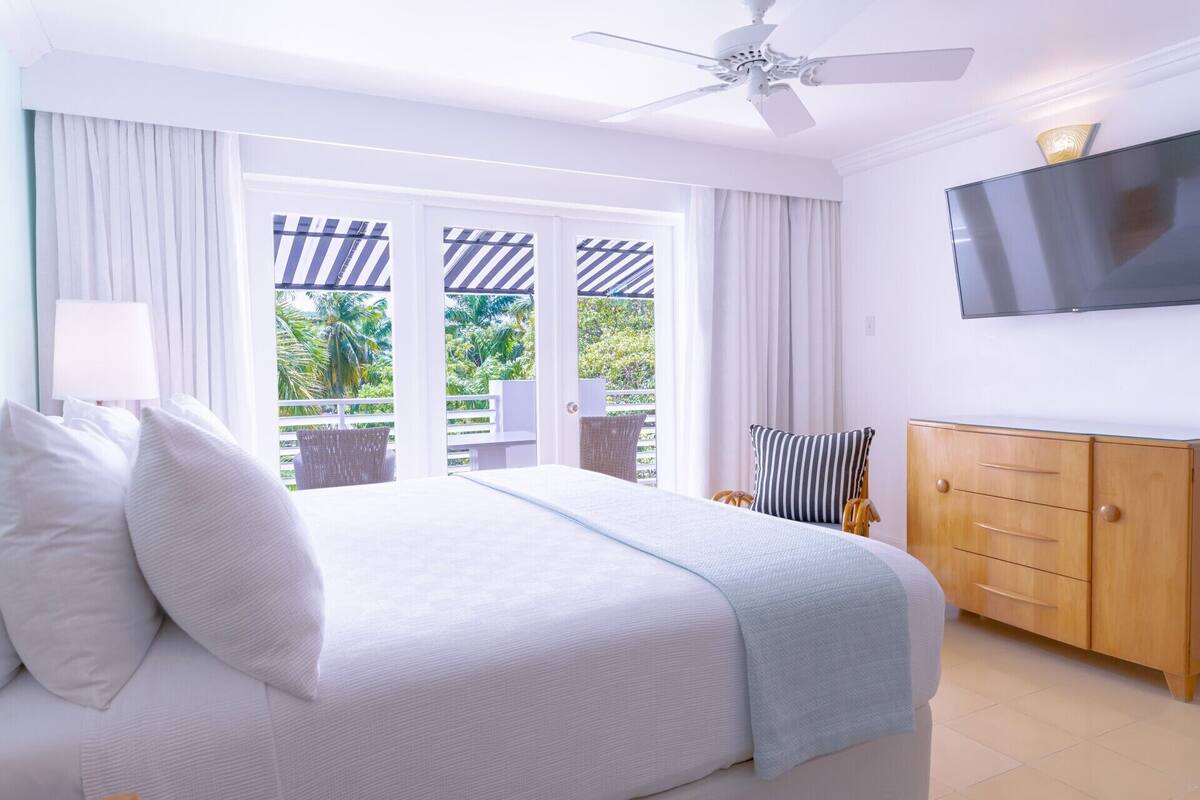 Quarto de hotel com cama, televisão presa na parede, armário de madeira e varanda com cortinas.