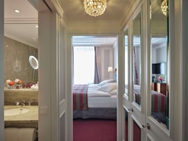 Quarto de hotel com uma cama grande, banheiro com pia e espelho, e vista da cidade pela janela.