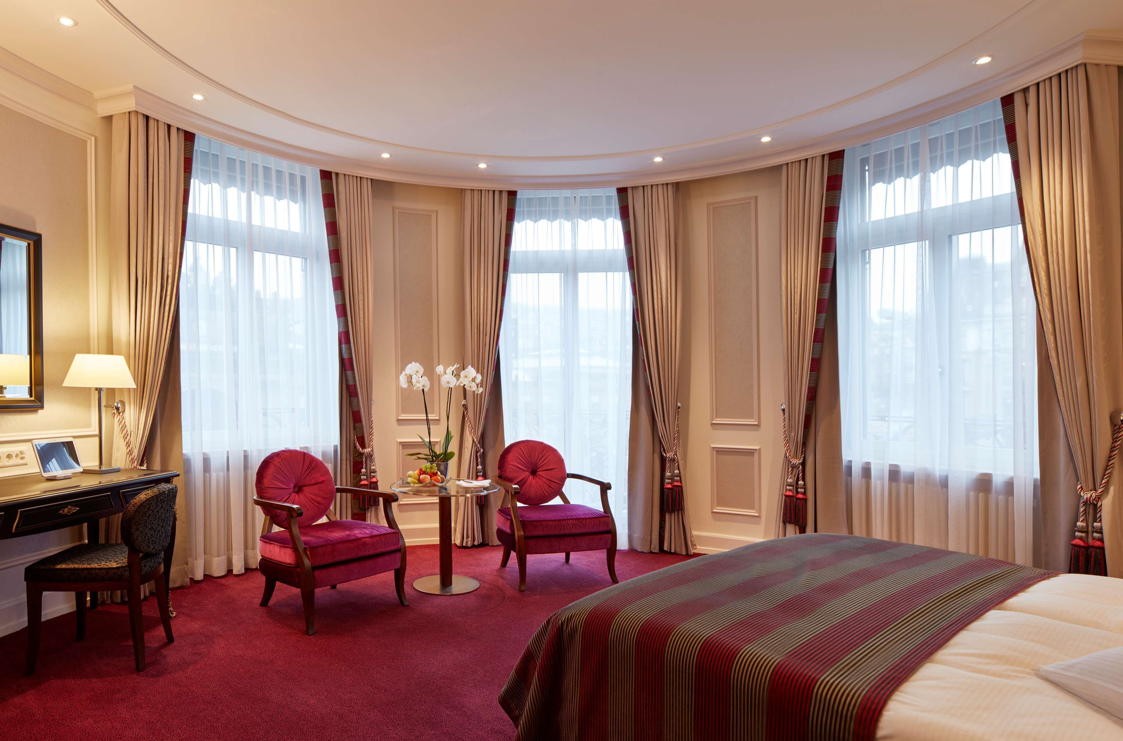 Quarto de hotel com uma cama grande, duas poltronas, uma escrivaninha com abajur e uma mesinha com flores.