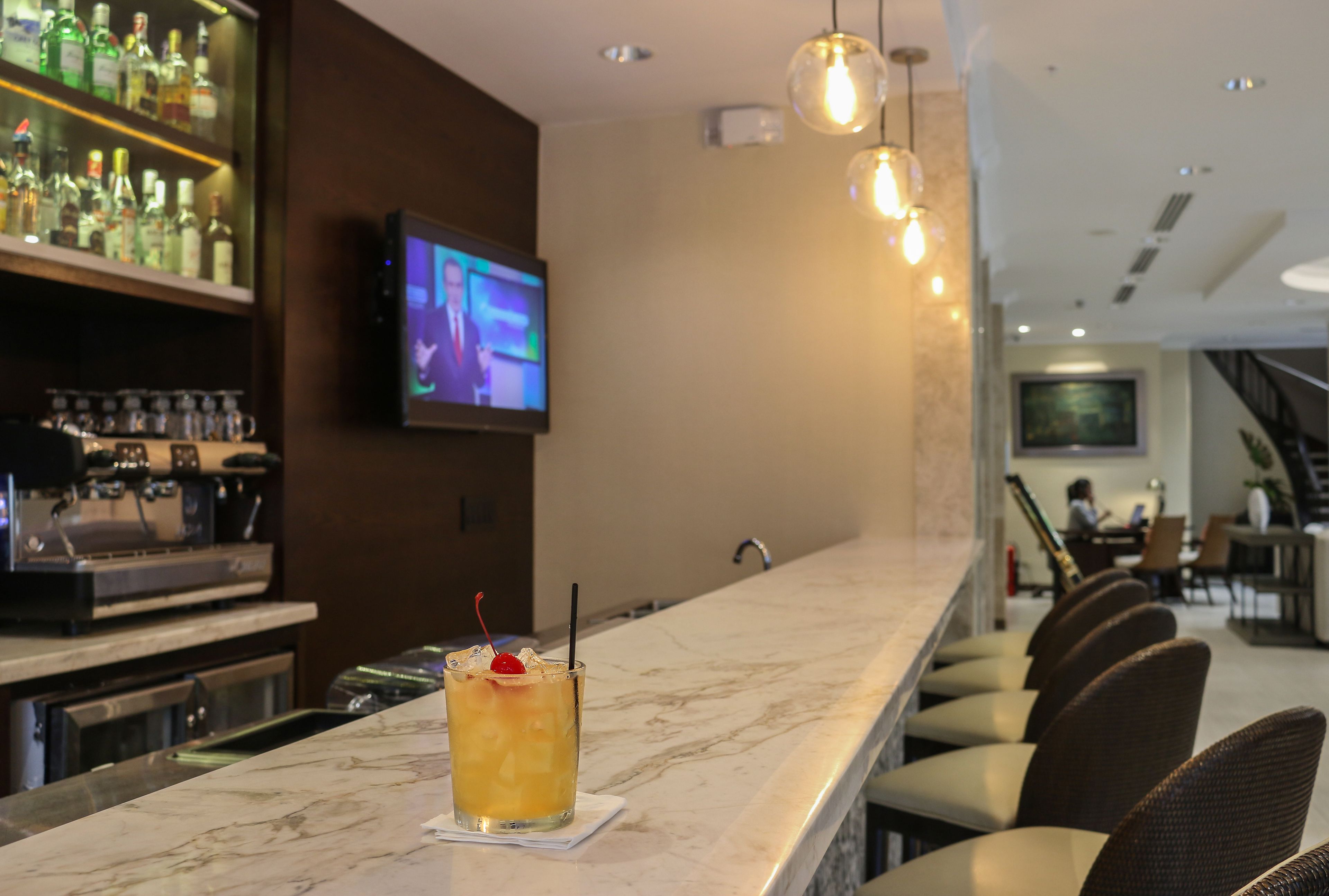 Uma área de bar com uma bebida no balcão, uma televisão fixada na parede e uma escada ao fundo.