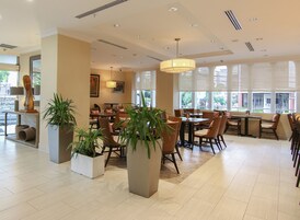 Uma sala de jantar ampla, com várias mesas e cadeiras, janelas grandes e plantas em vasos.