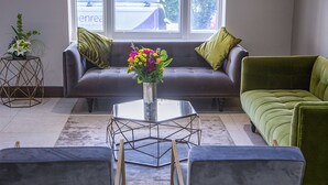 Uma sala de estar moderna com um sofá cinzento, poltronas verdes e uma mesa de centro de vidro com um arranjo floral.