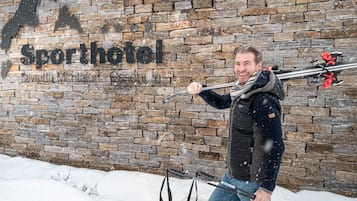 Uma pessoa segurando bastões de esqui em frente a um muro de tijolos com os dizeres "Sporthotel" e "Silvretta Montan Gaschuhi".