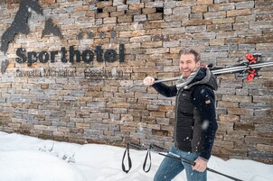 Uma pessoa segurando bastões de esqui em frente a um muro de tijolos com os dizeres "Sporthotel" e "Silvretta Montan Gaschuhi".