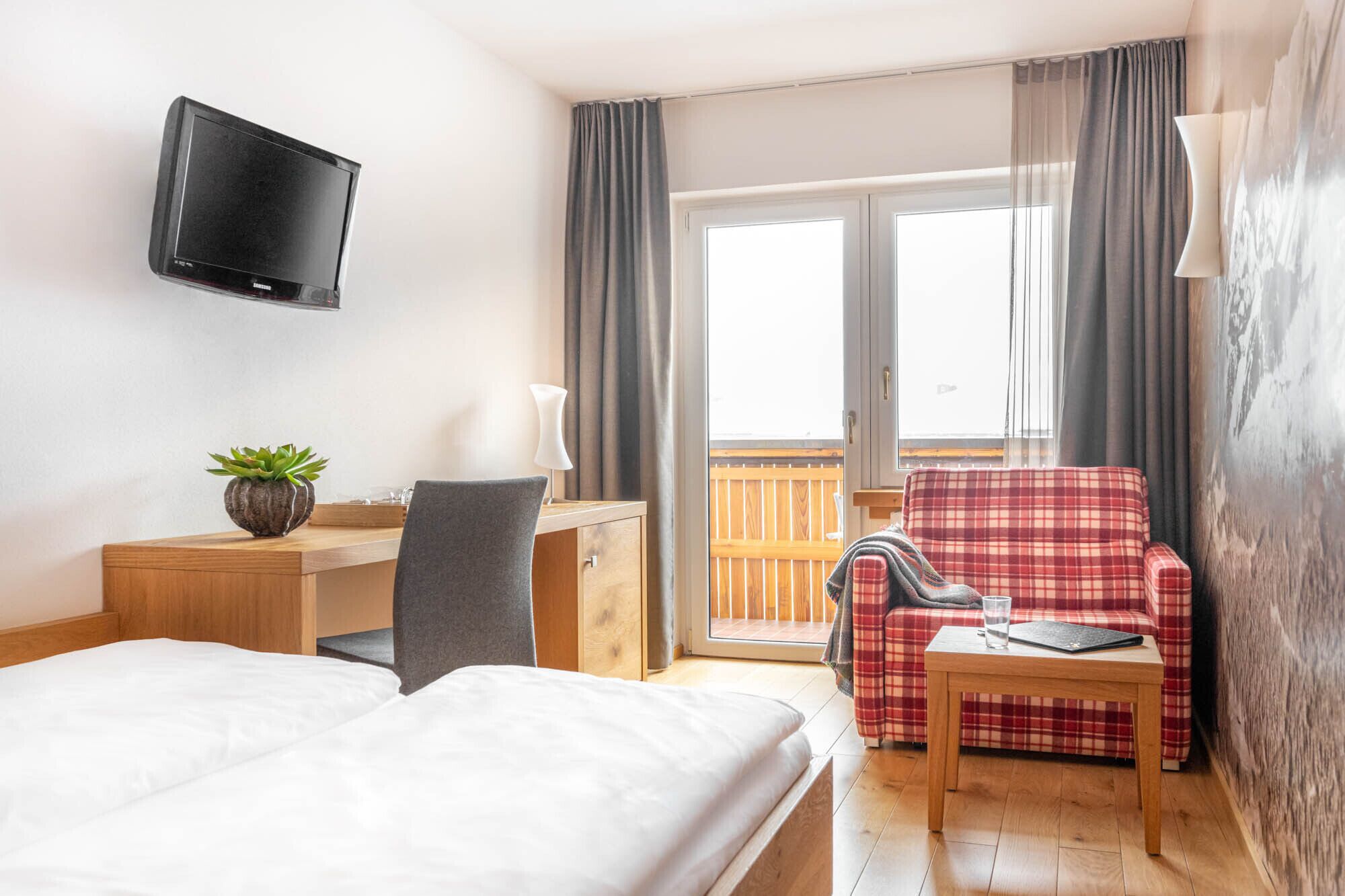 Quarto de hotel com cama, escrivaninha com cadeira, sofá xadrez vermelho, mesinha, televisão fixada na parede e varanda com cortinas.